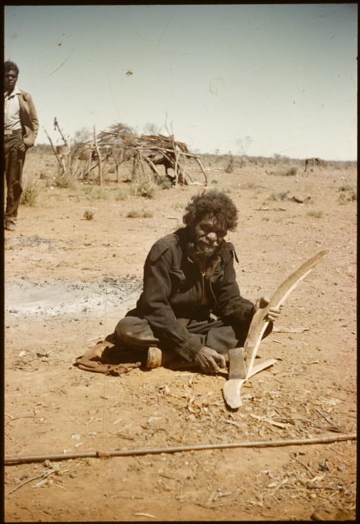Aboriginal man seated on the ground in the desert making a boomerang ...