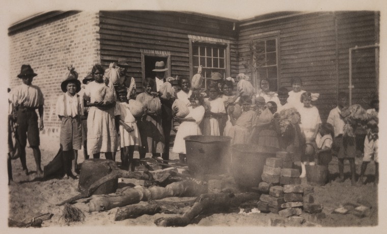 Aboriginal people holding puddings behind large pots on a campfire ...
